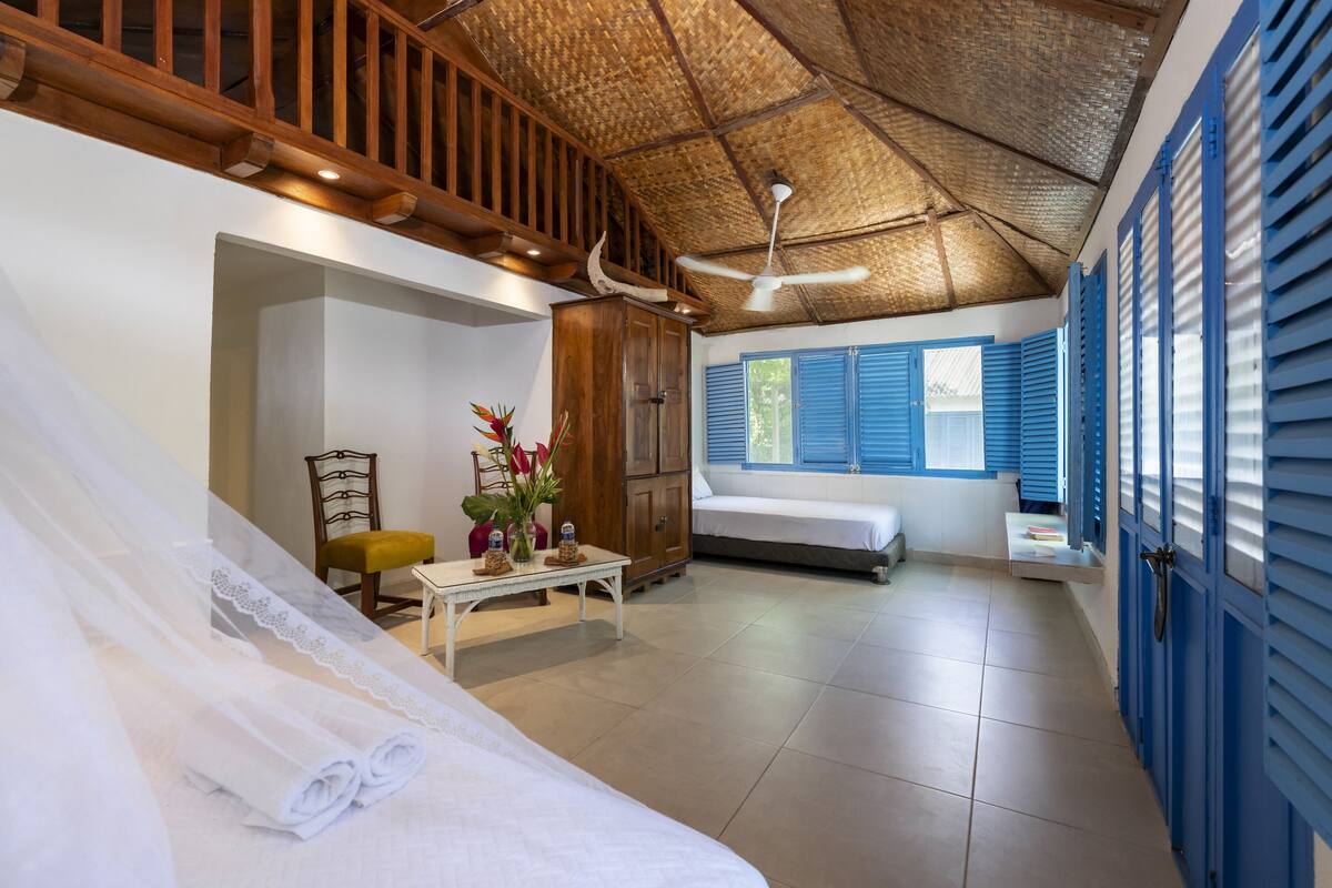 Quarto com cama, um banco de madeira, mesa com flores, ventilador de teto e janela com persianas azuis.