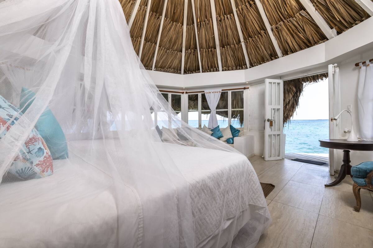 Quarto com cama coberta por um dossel branco, vista para o oceano e telhado de palha.