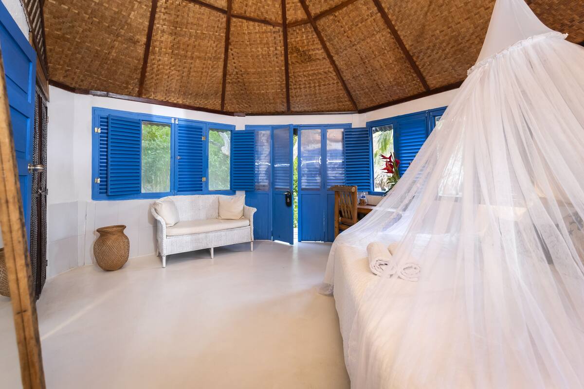 Quarto com cama, sofá branco, cadeira de vime e uma grande janela com persianas azuis.