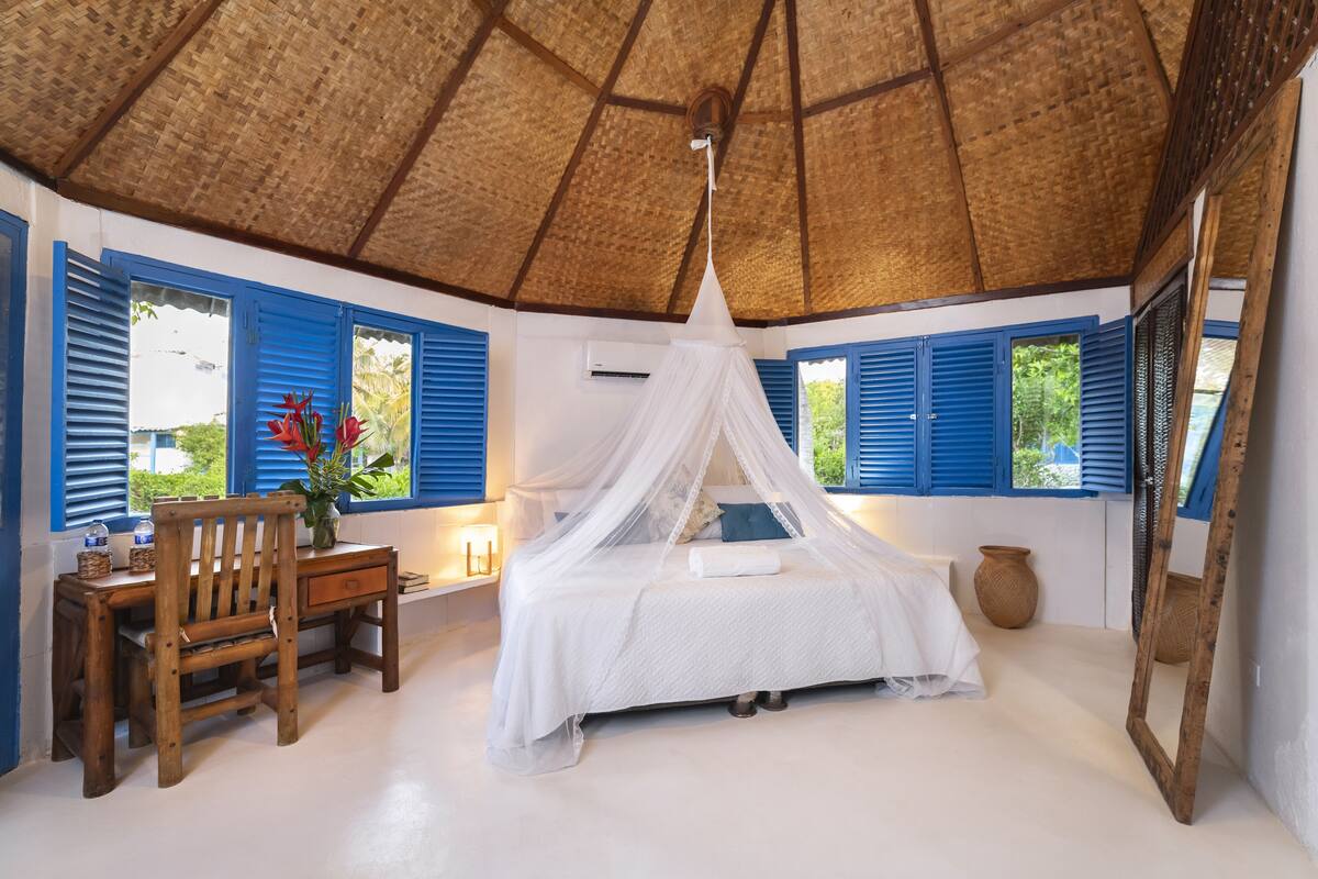 Quarto com cama sob telhado de palha, persianas azuis, uma cadeira de madeira, mesa de cabeceira e um vaso com flores.
