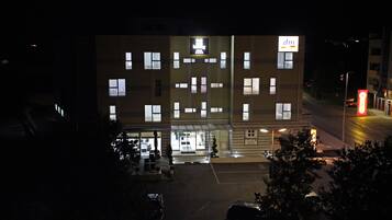 Um hotel com vários andares, entrada central, estacionamento e uma placa visível com 'dm' na fachada.
