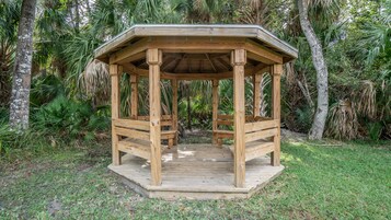 Um gazebo de madeira com telhado hexagonal e laterais abertas, rodeado por vegetação exuberante.