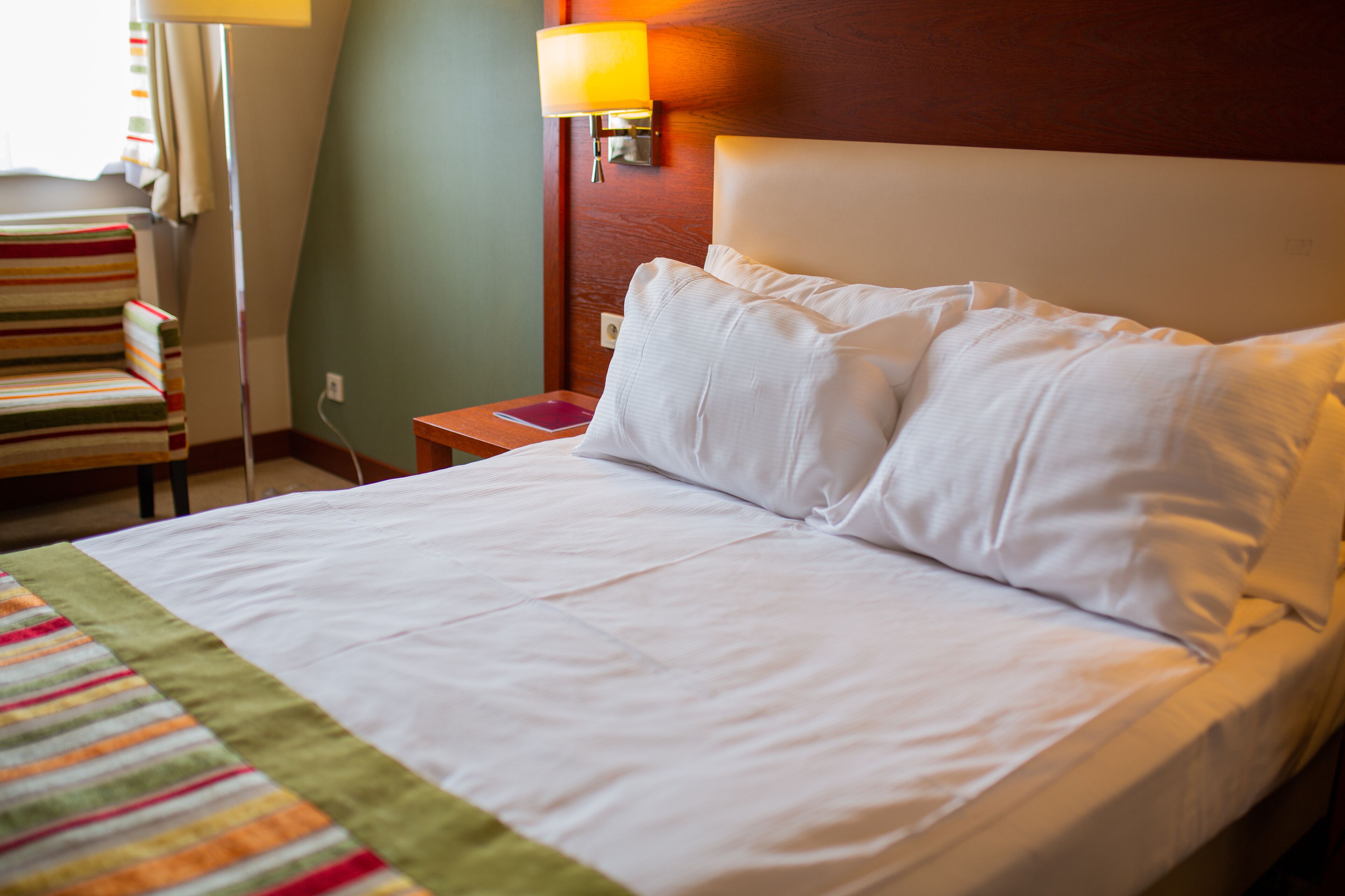 Cama bem arrumada com lençóis brancos, uma cadeira listrada e uma mesa de cabeceira com abajur.