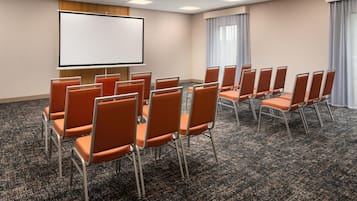 Sala de conferências com fileiras de cadeiras laranja, uma grande tela de projeção branca e piso coberto de carpete.