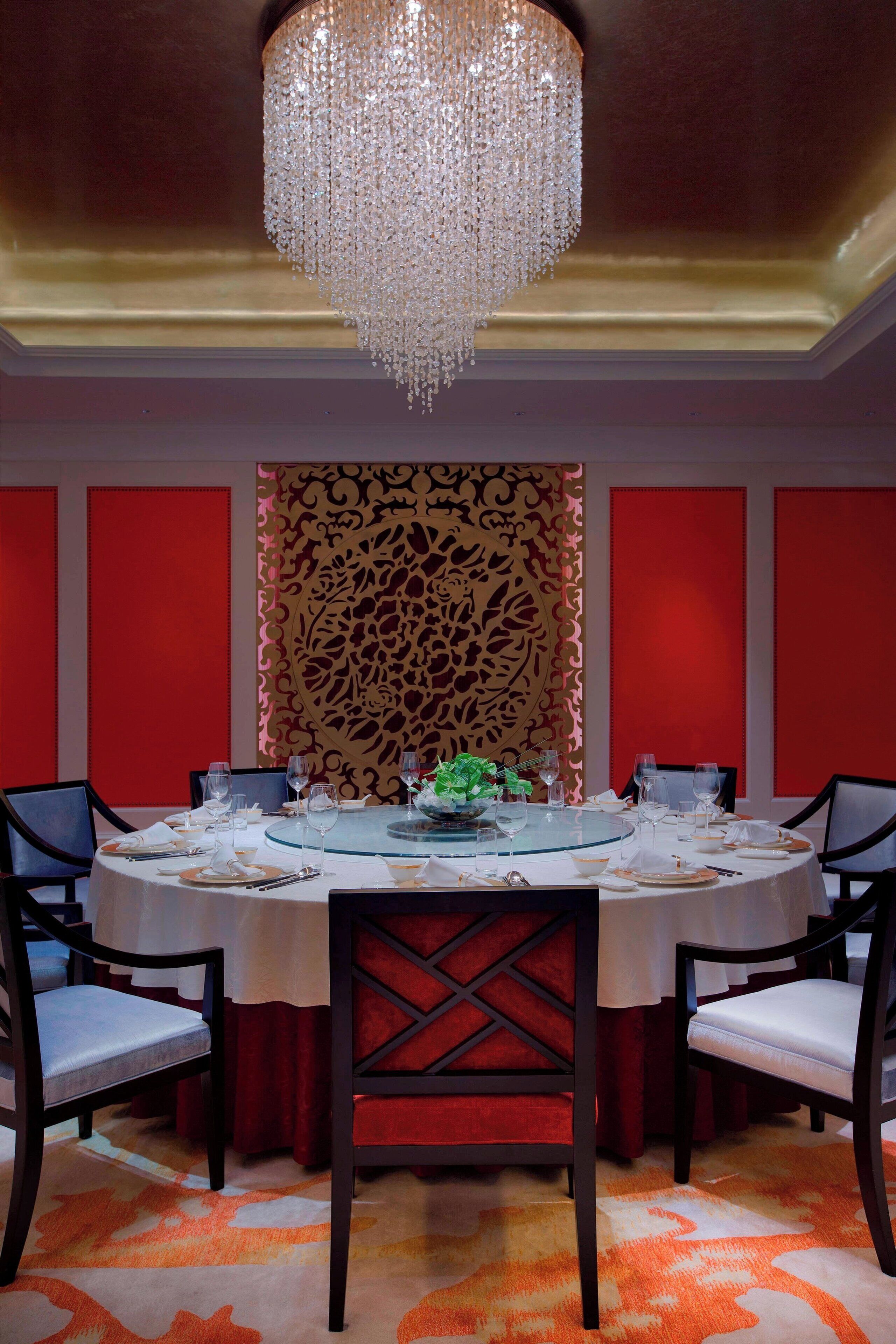 Uma área de refeições com uma mesa redonda posta para quatro pessoas, rodeada por cadeiras. Um candelabro pendurado no teto, e as paredes apresentam um design com padrão em vermelho e dourado.