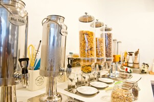 Disposição de buffet com diversos distribuidores de alimentos, incluindo um dispensador de leite quente e um dispensador de cereais.