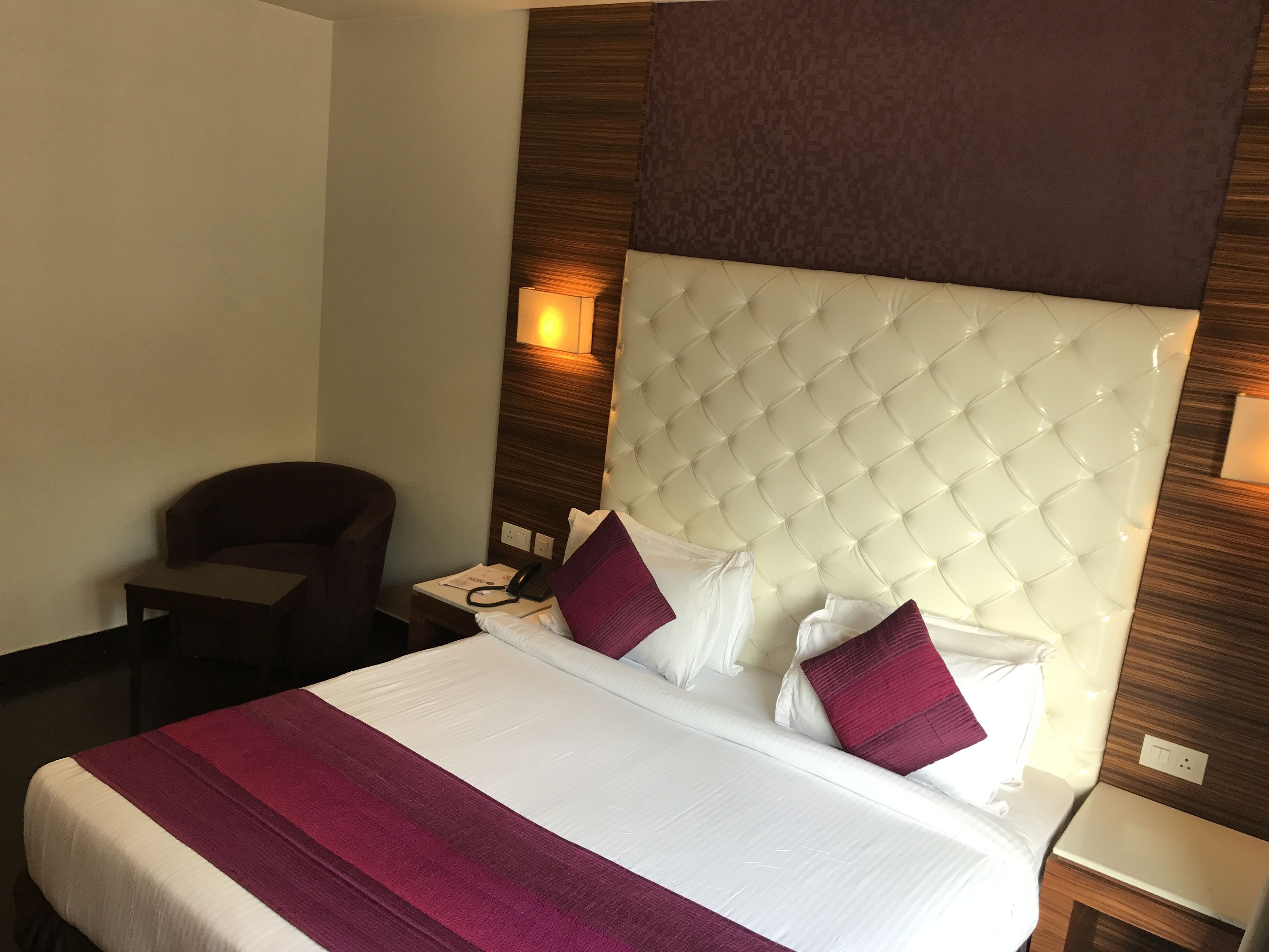 Quarto de hotel com uma cama grande, cabeceira de madeira, mesa de cabeceira, uma cadeira e luminária fixada na parede.