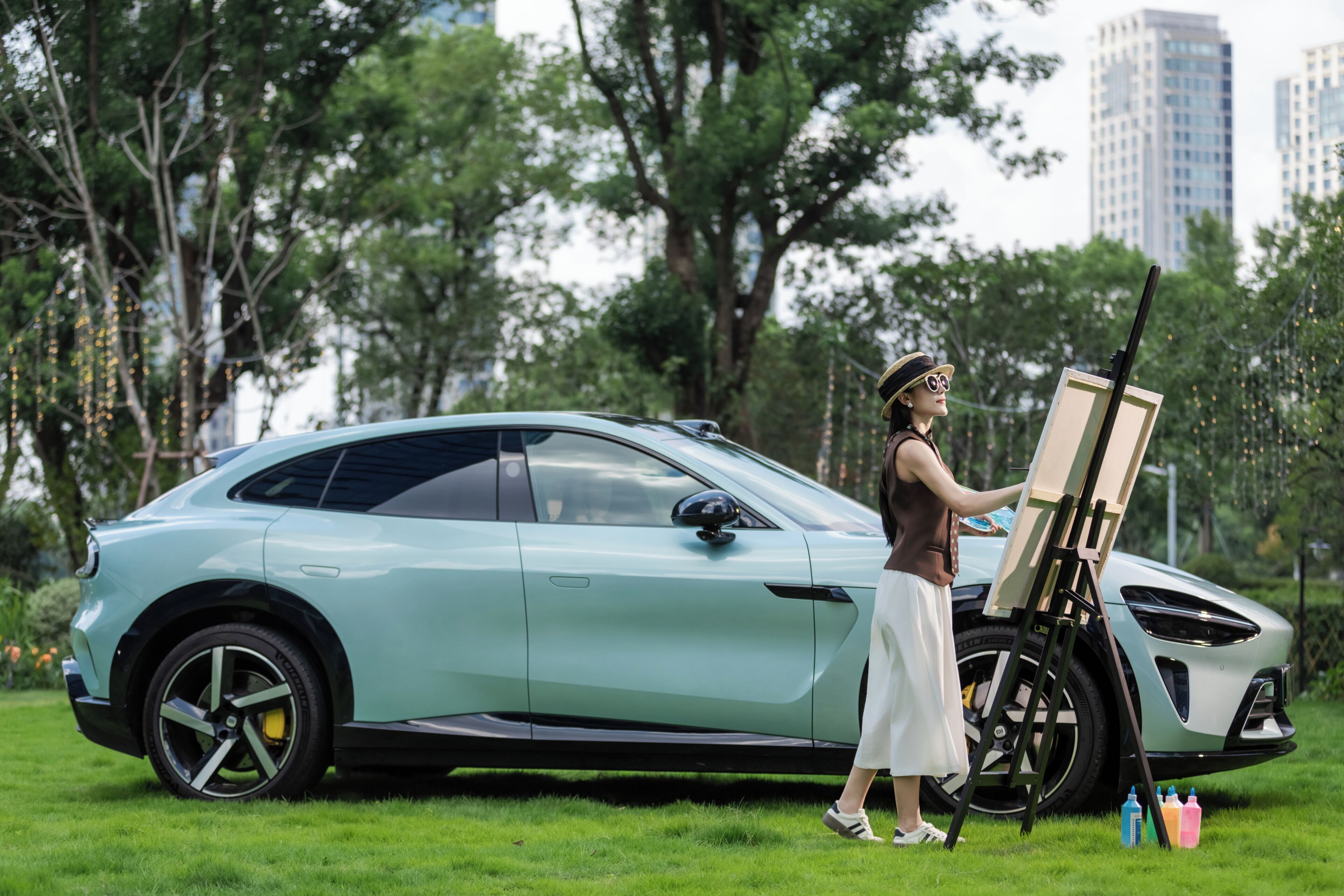 Uma pessoa pinta perto de um carro azul-claro numa área gramada, com árvores e edifícios ao fundo.
