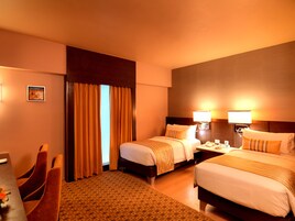 Quarto de hotel com duas camas, uma escrivaninha e uma cadeira. Há uma janela com cortinas, uma luminária na parede e uma mesinha com um telefone.