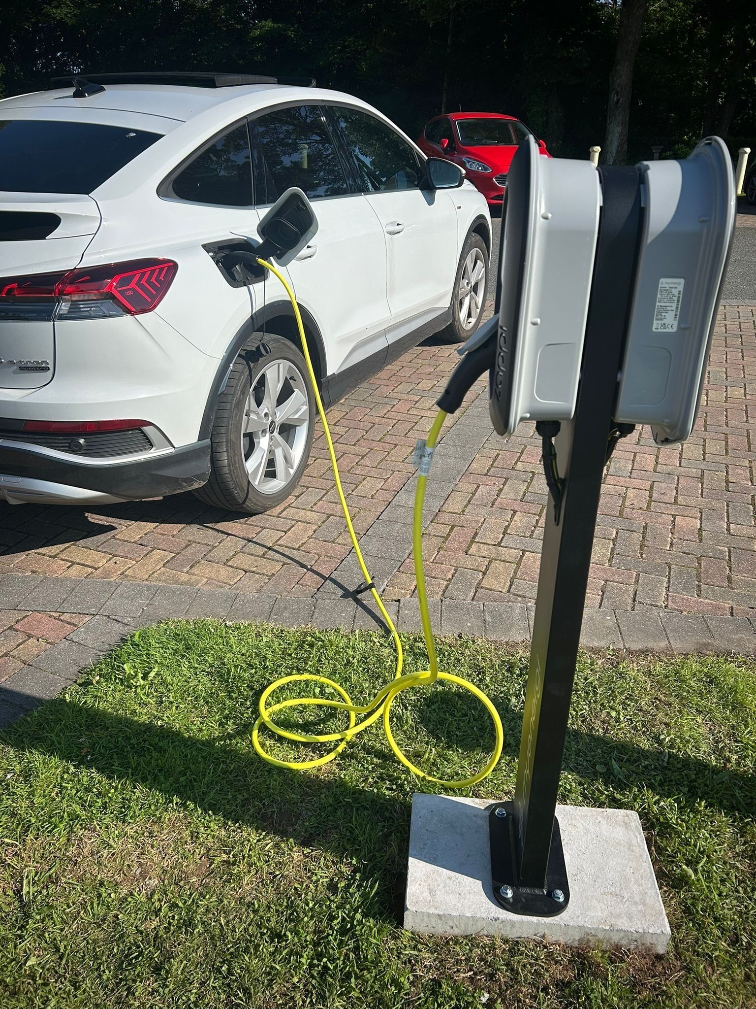 Um carro branco estacionado ao lado de uma estação de carregamento com um cabo amarelo conectado.