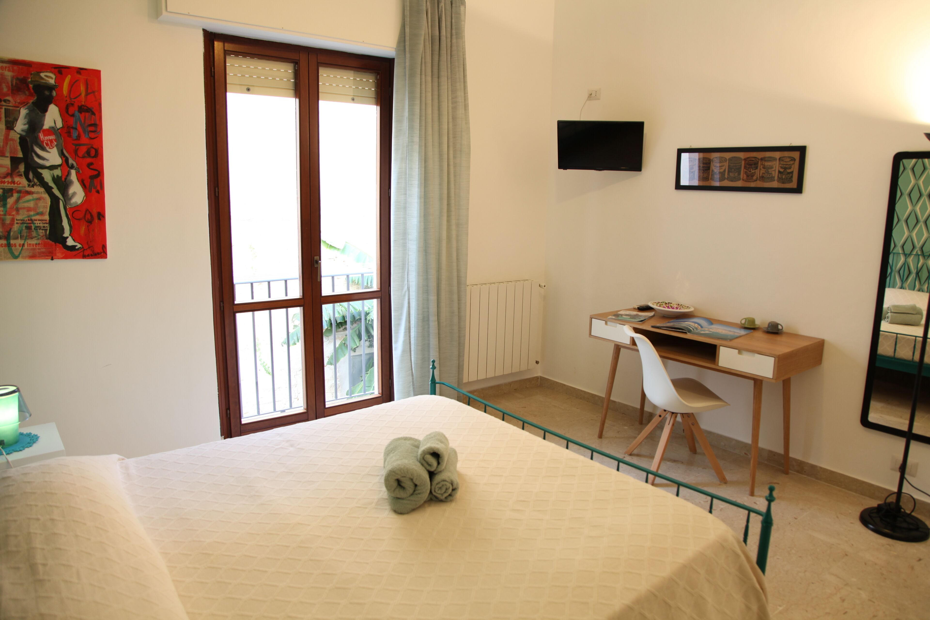 Quarto com cama, escrivaninha, cadeira, janela com cortinas e televisão na parede.