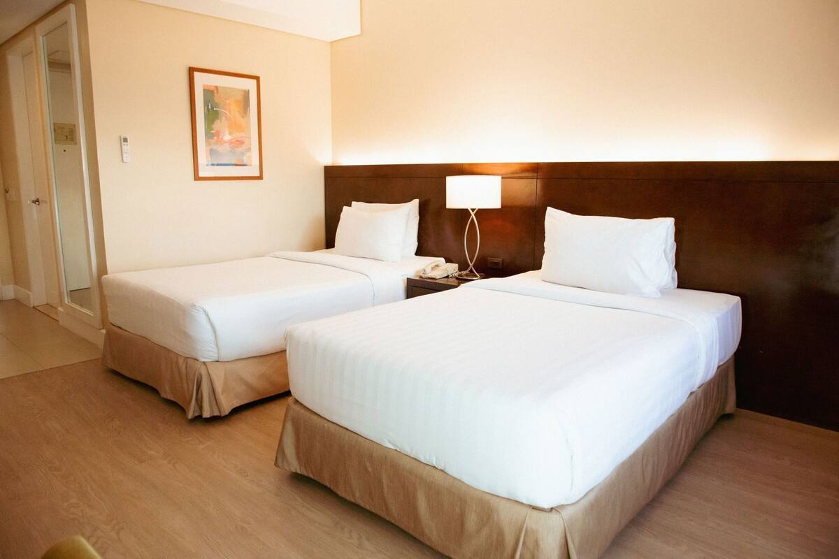 Quarto de hotel com duas camas, uma mesa de cabeceira com abajur e um quadro na parede.