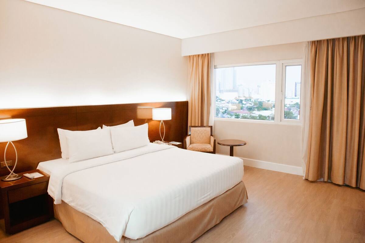 Quarto de hotel com uma cama grande, mesa de cabeceira, uma cadeira, uma pequena mesa e uma janela com cortinas.