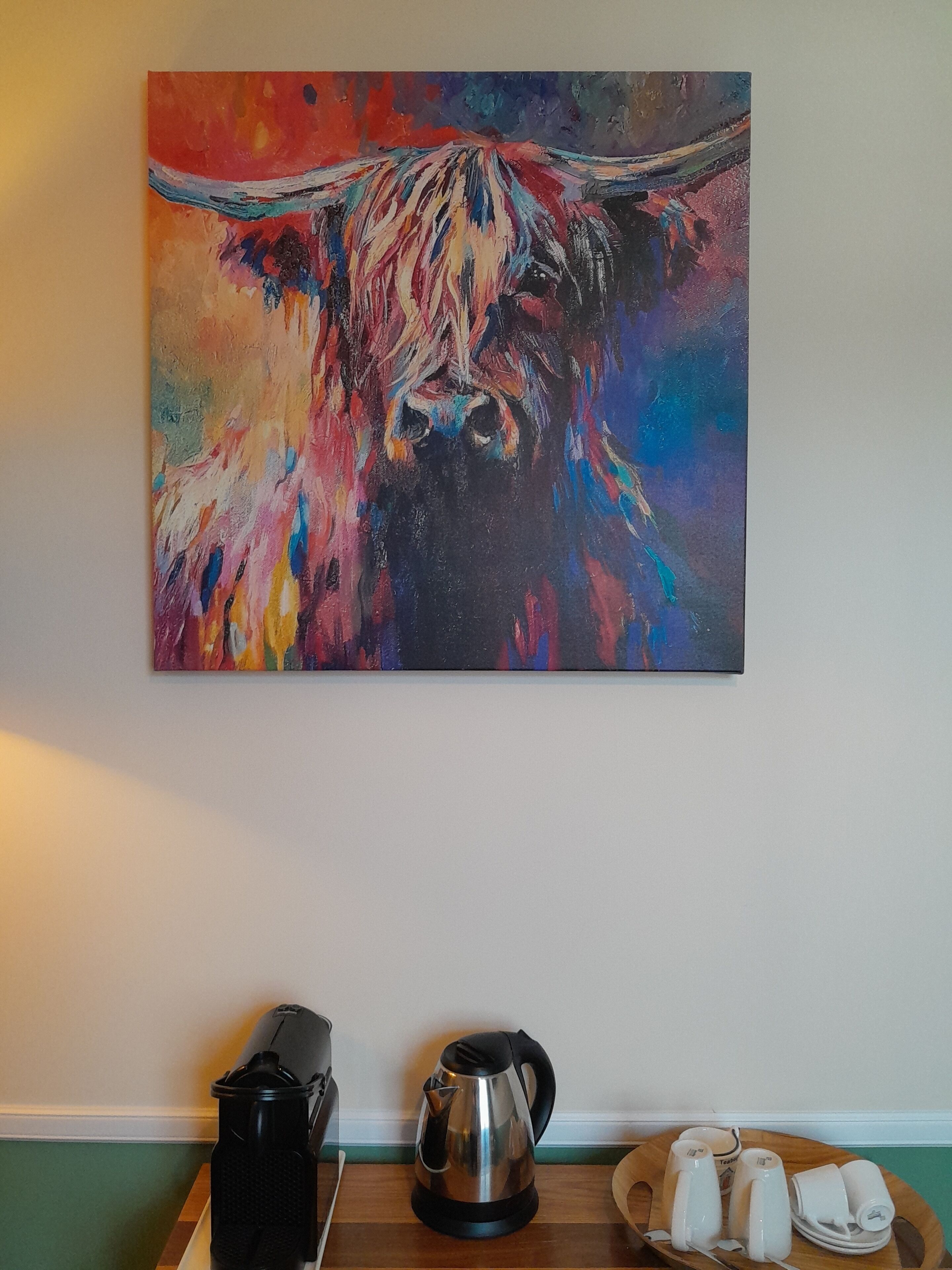 Pintura de cabeça de touro com um fundo colorido, uma chaleira, uma cafeteira e uma bandeja com chávenas e um carregador sobre uma mesa.