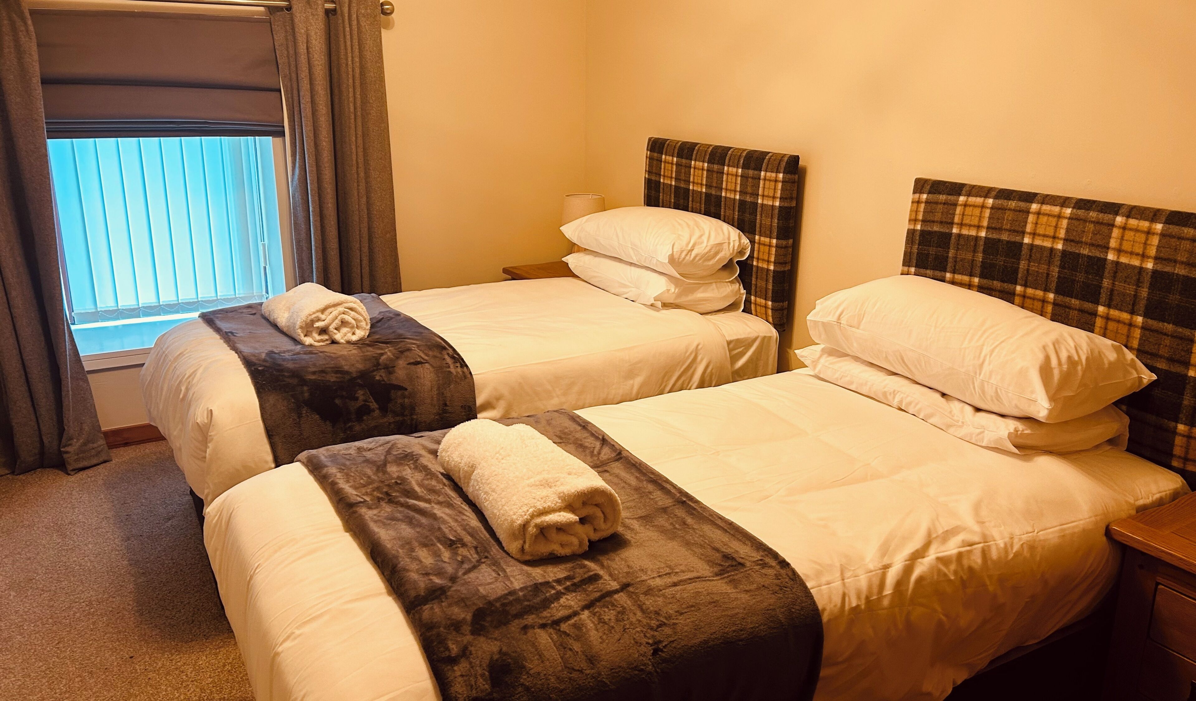 Quarto de hotel com duas camas, uma janela com cortinas e uma mesa de cabeceira com abajur.