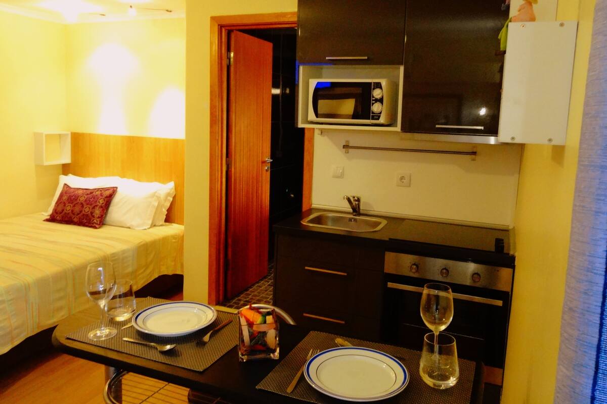 Quarto de hotel compacto com cama, pequeno espaço de cozinha e área de refeições com mesa posta para dois.