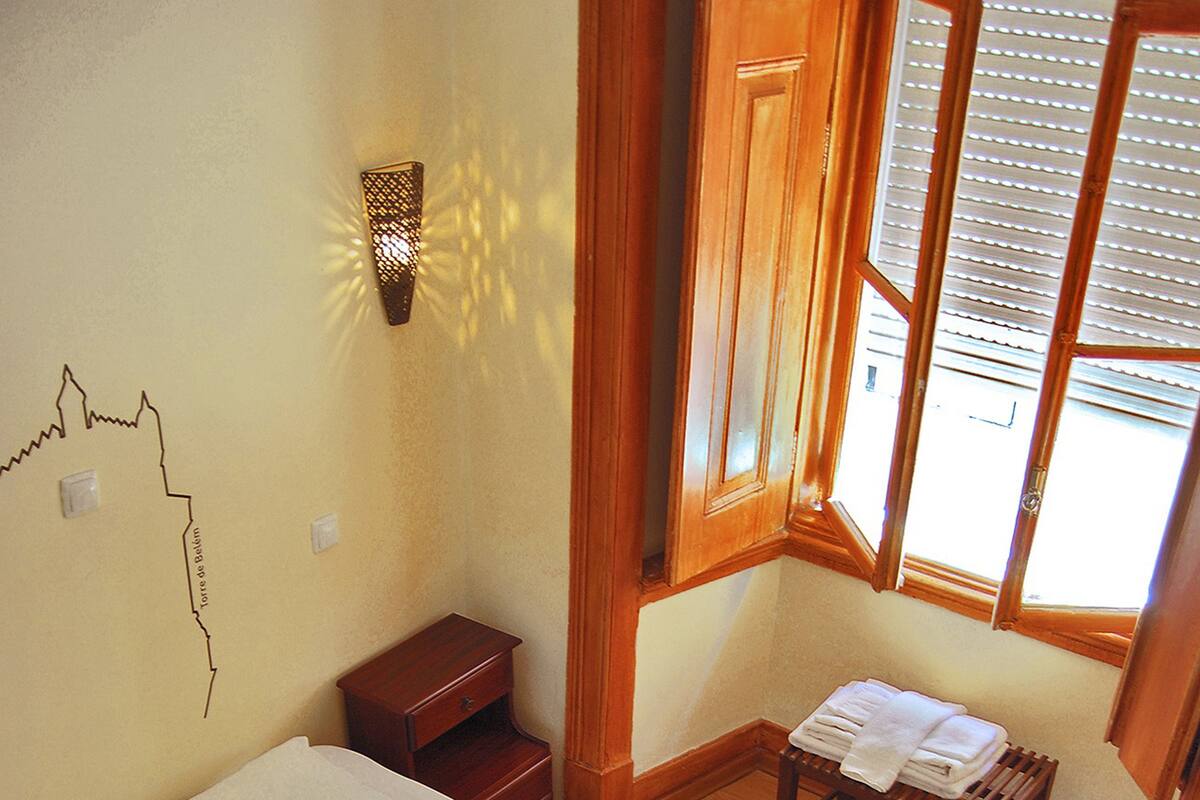 Quarto com cama, mesa de cabeceira, pequena mesa de madeira com toalhas e luminária na parede.