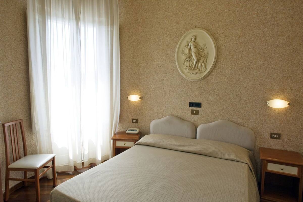 Quarto de hotel com cama, poltrona, mesa de cabeceira, luminária na parede e um prato decorativo.