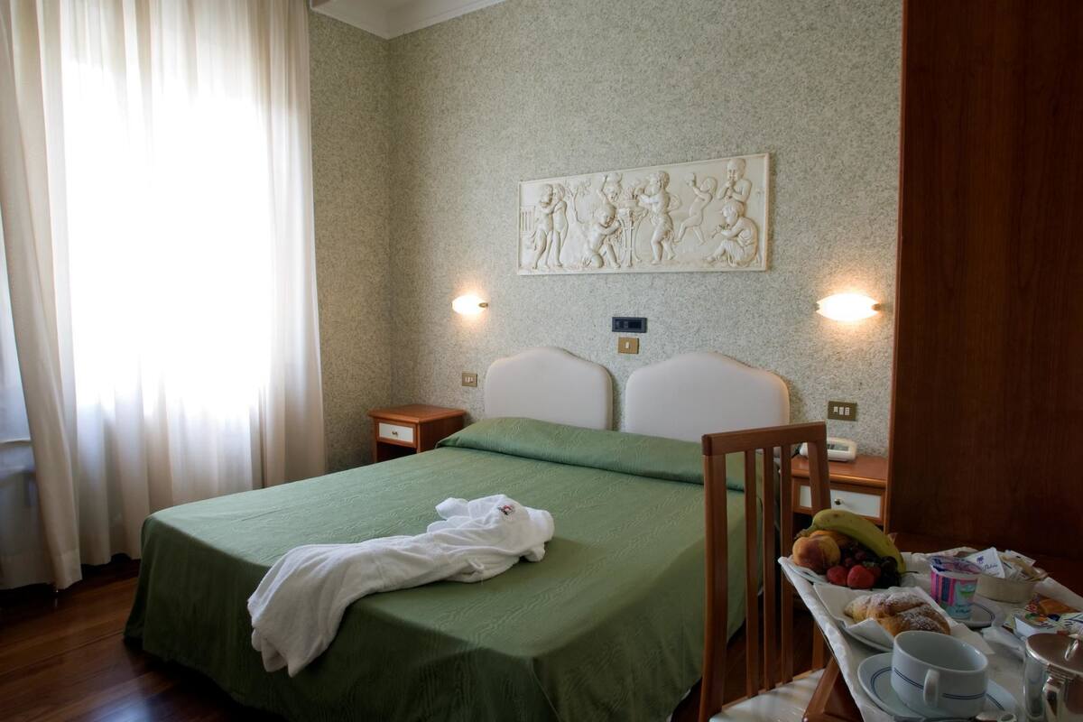 Quarto de hotel com cama verde, toalha branca, bandeja com itens de café da manhã e parede com escultura em relevo.