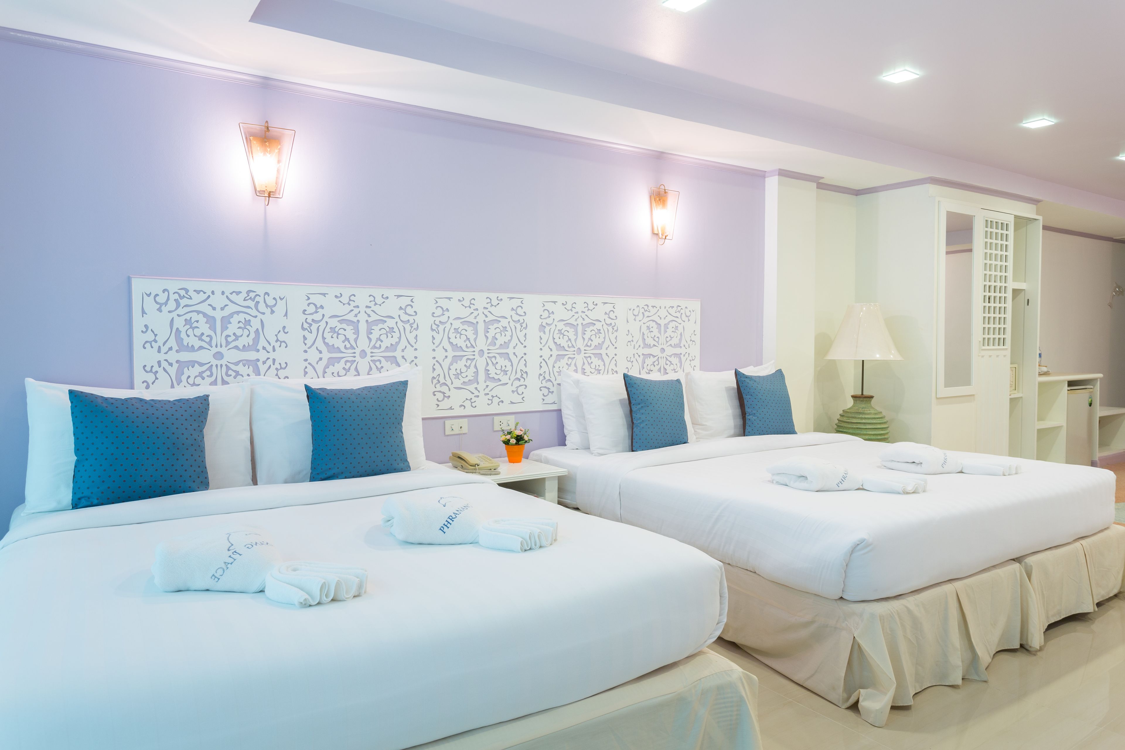 Quarto de hotel com duas camas, roupa de cama branca, cabeceira decorativa, arandelas, mesa de cabeceira e abajur.