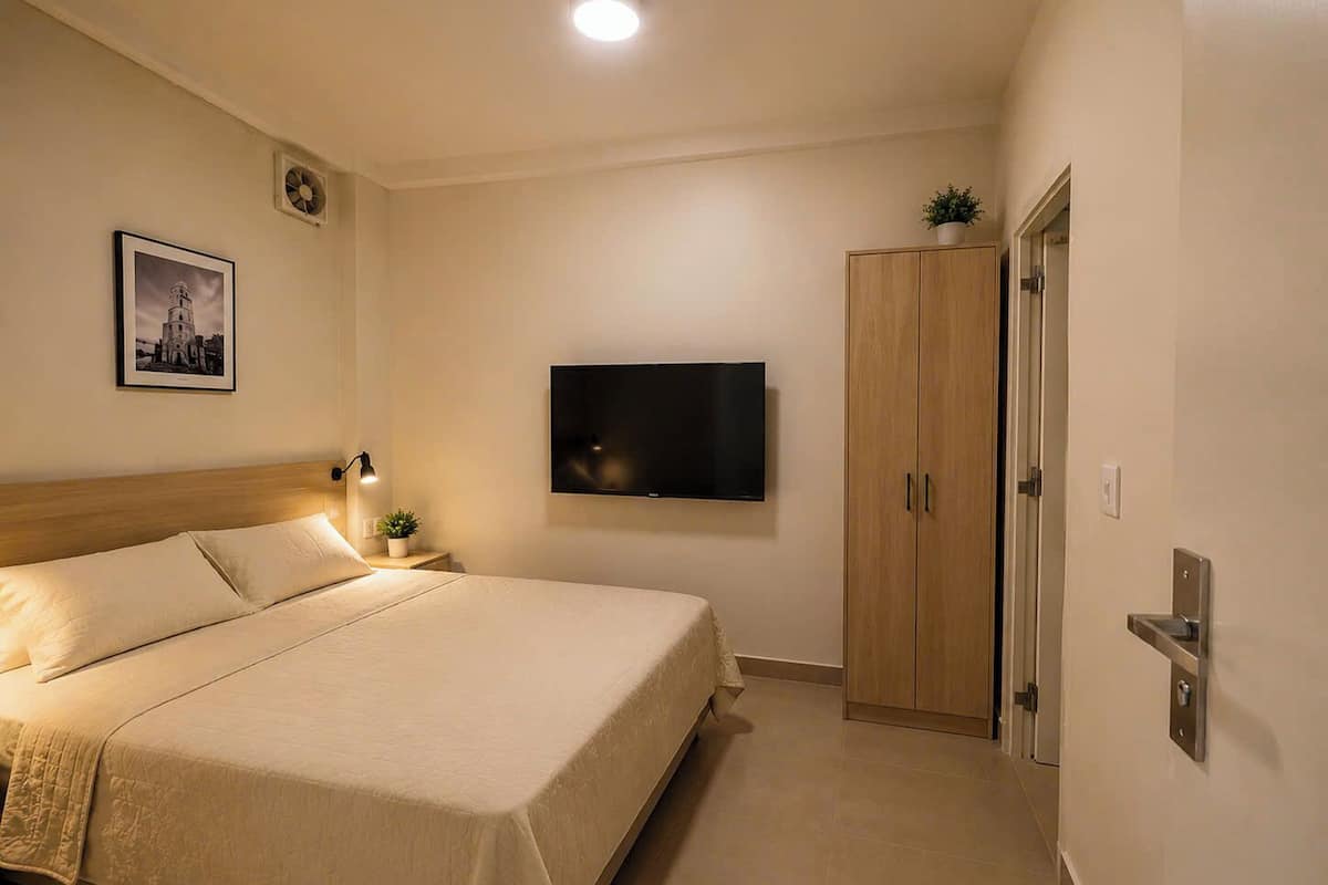 Quarto com cama, televisão, guarda-roupa e uma pequena mesa com abajur.