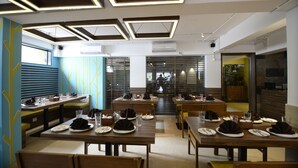 Restaurante com várias mesas preparadas para refeições, apresentando design de interiores moderno com luminárias geométricas no teto e vista para a cozinha ao fundo.