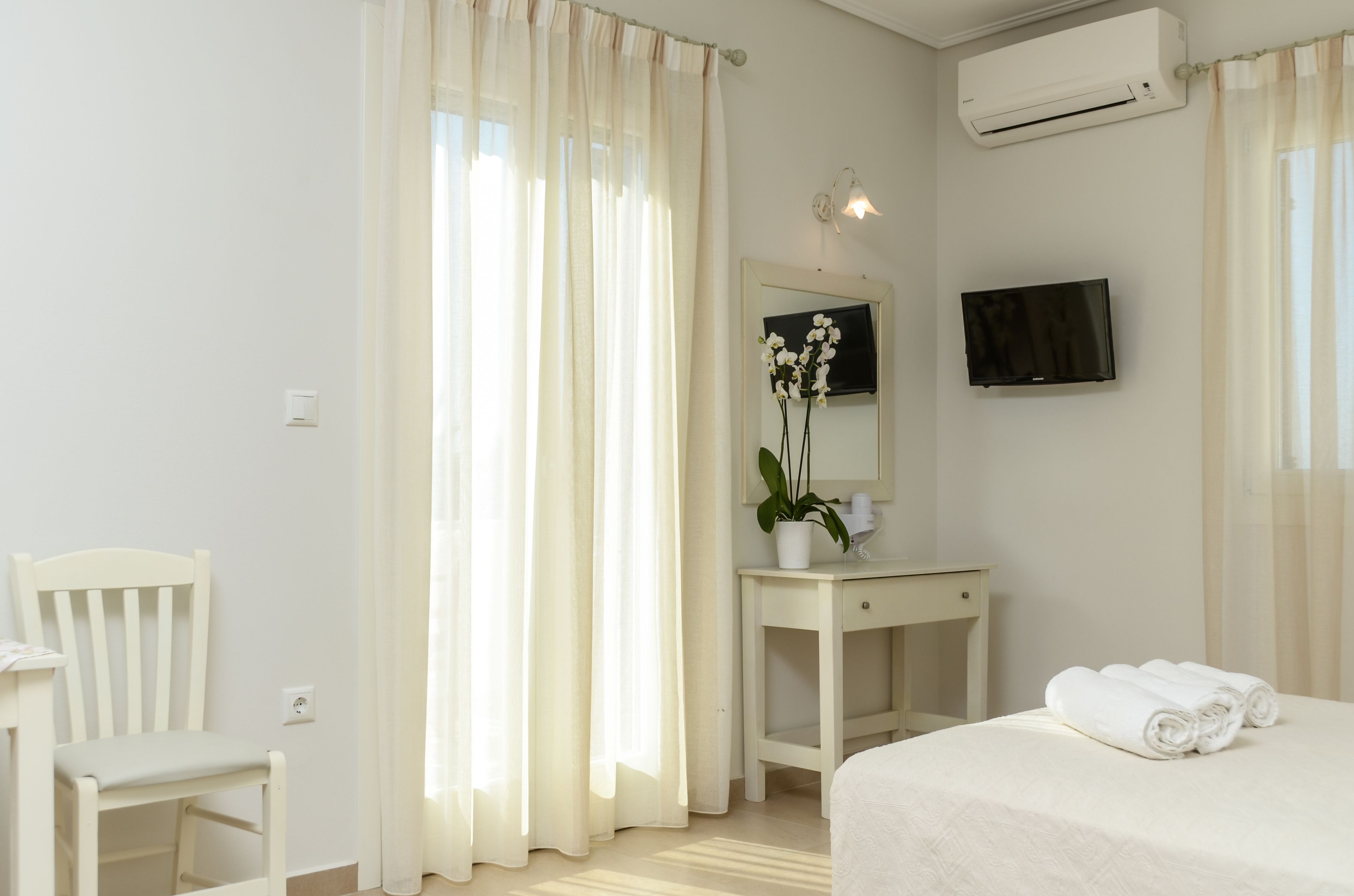 Quarto de hotel com cama, cadeira, escrivaninha, espelho, televisão e cortinas.