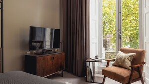 Quarto de hotel com cama, televisão sobre um móvel de madeira, poltrona com almofada e vista para prédios através de grandes janelas.