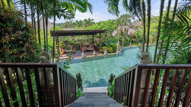 Uma área de piscina com uma escadaria de pedra que desce até ela, rodeada por vegetação exuberante e palmeiras.