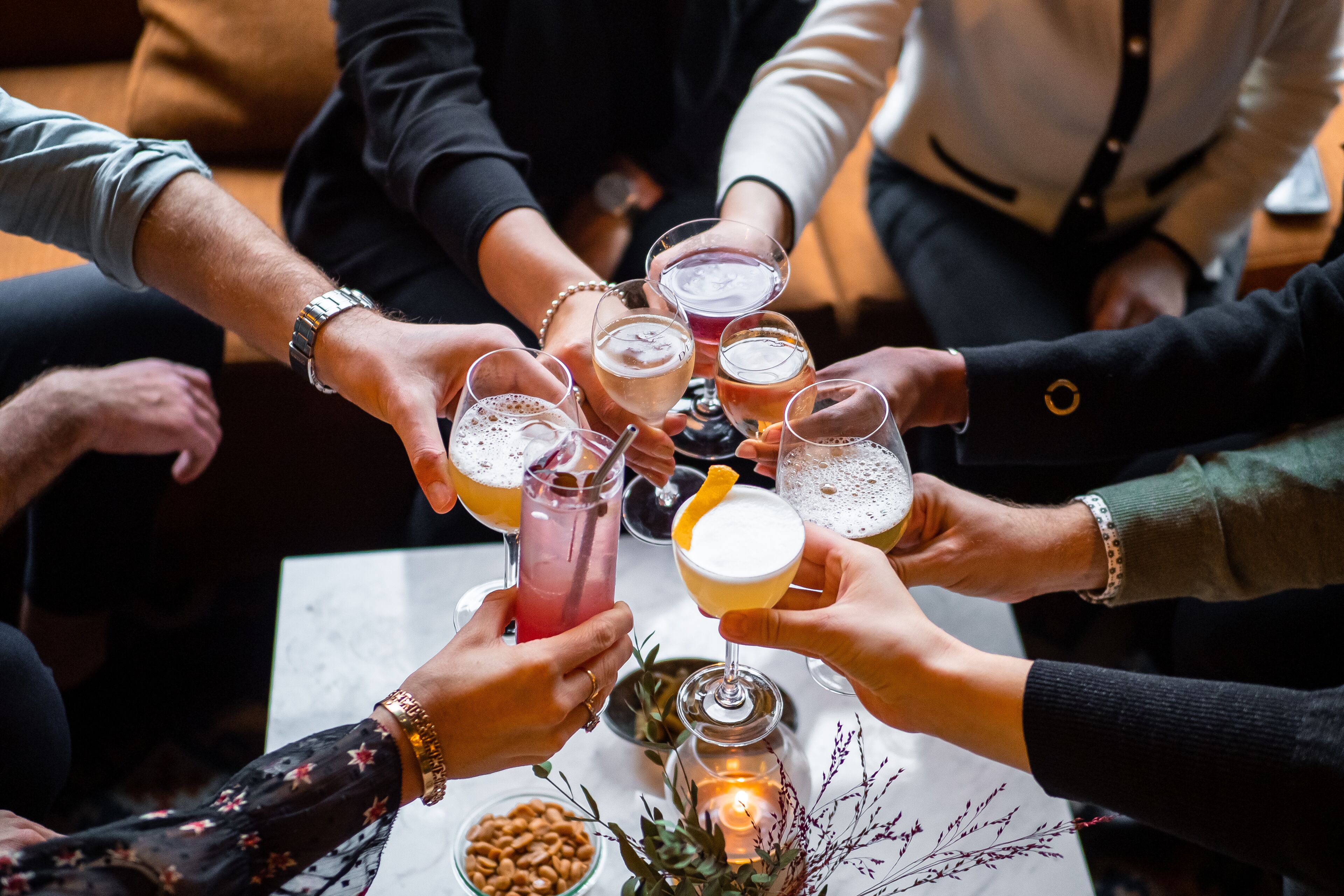 Um grupo de pessoas brindando com diferentes bebidas em uma mesa com vela e plantas decorativas.