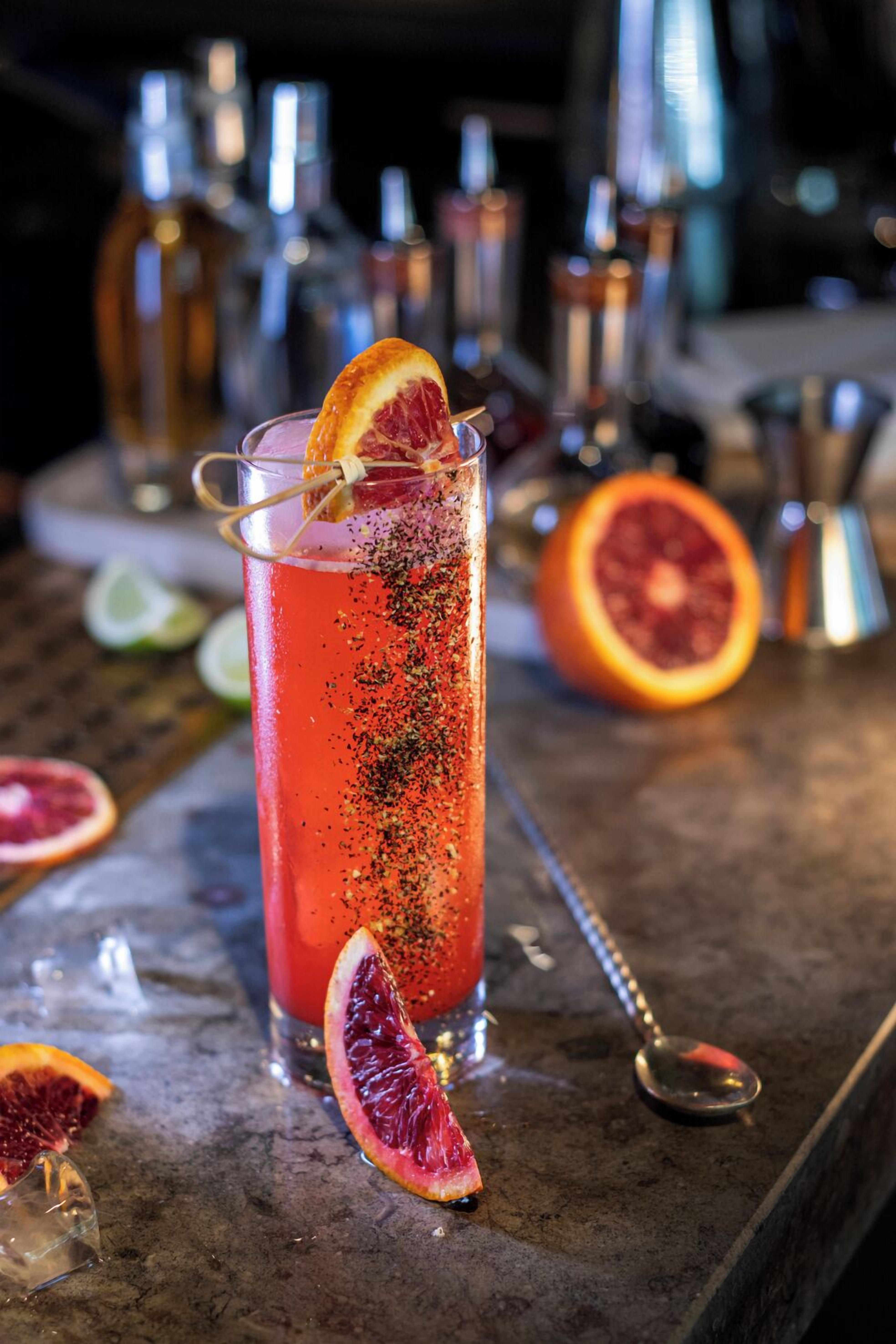 Um copo alto com uma bebida vermelha, decorado com uma fatia de laranja sanguínea e pimenta do reino, colocado sobre um balcão de bar, ao lado de uma colher e meia laranja sanguínea.