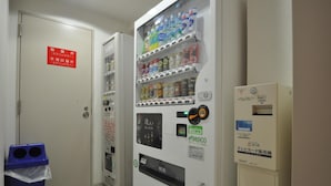 Máquina de vendas com diversos snacks e bebidas, uma lixeira e uma porta com placa vermelha.