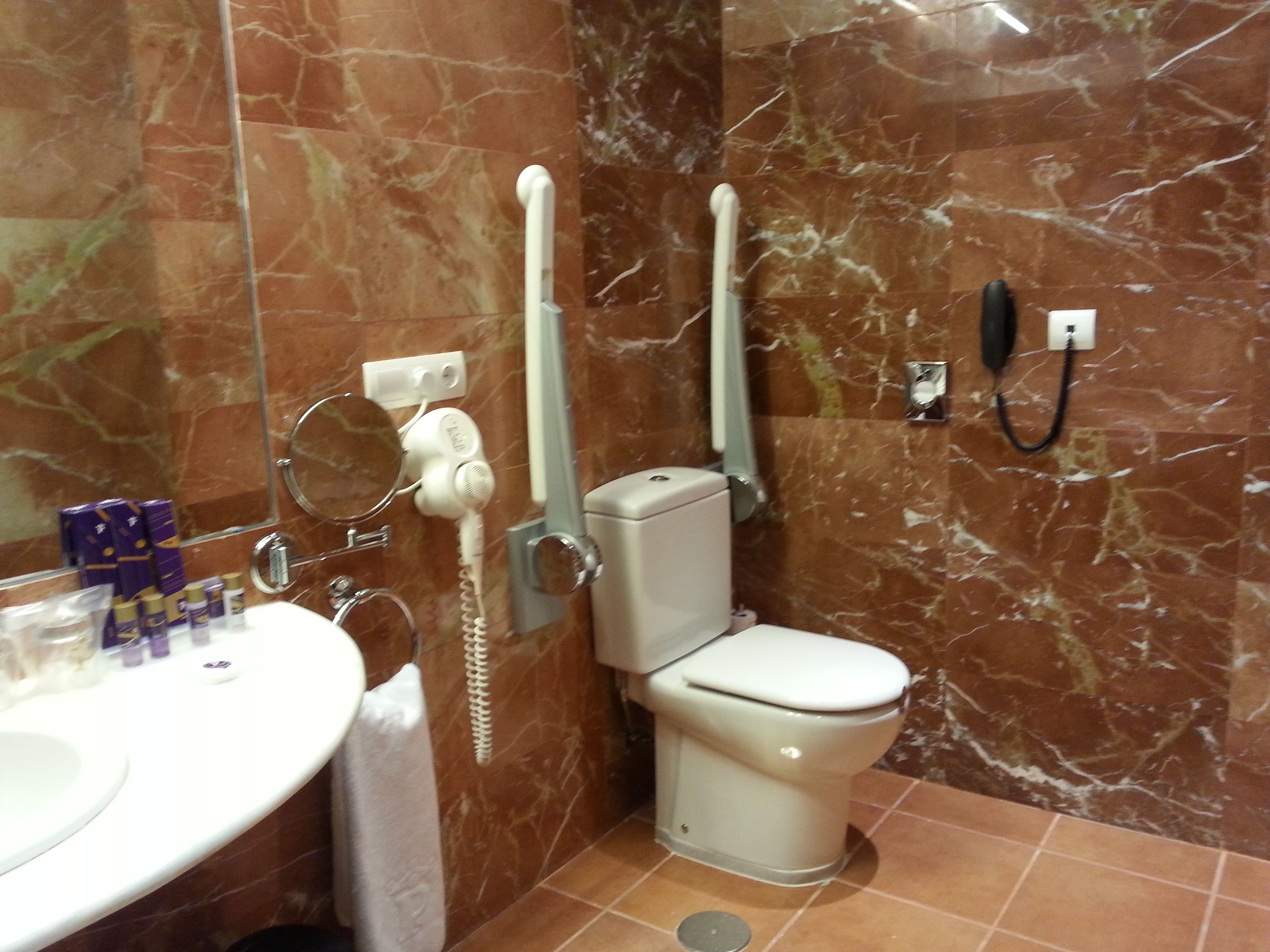 Casa de banho com lavatório branco, espelho e sanita. Há produtos de higiene no lavatório e uma toalha pendurada na parede.