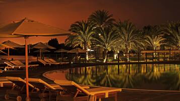 Área de piscina à noite com espreguiçadeiras e guarda-sóis, cercada por palmeiras e uma piscina calma que reflete as luzes.