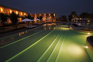 Piscina com espreguiçadeiras e guarda-sóis, cercada por palmeiras e um edifício com iluminação acolhedora.