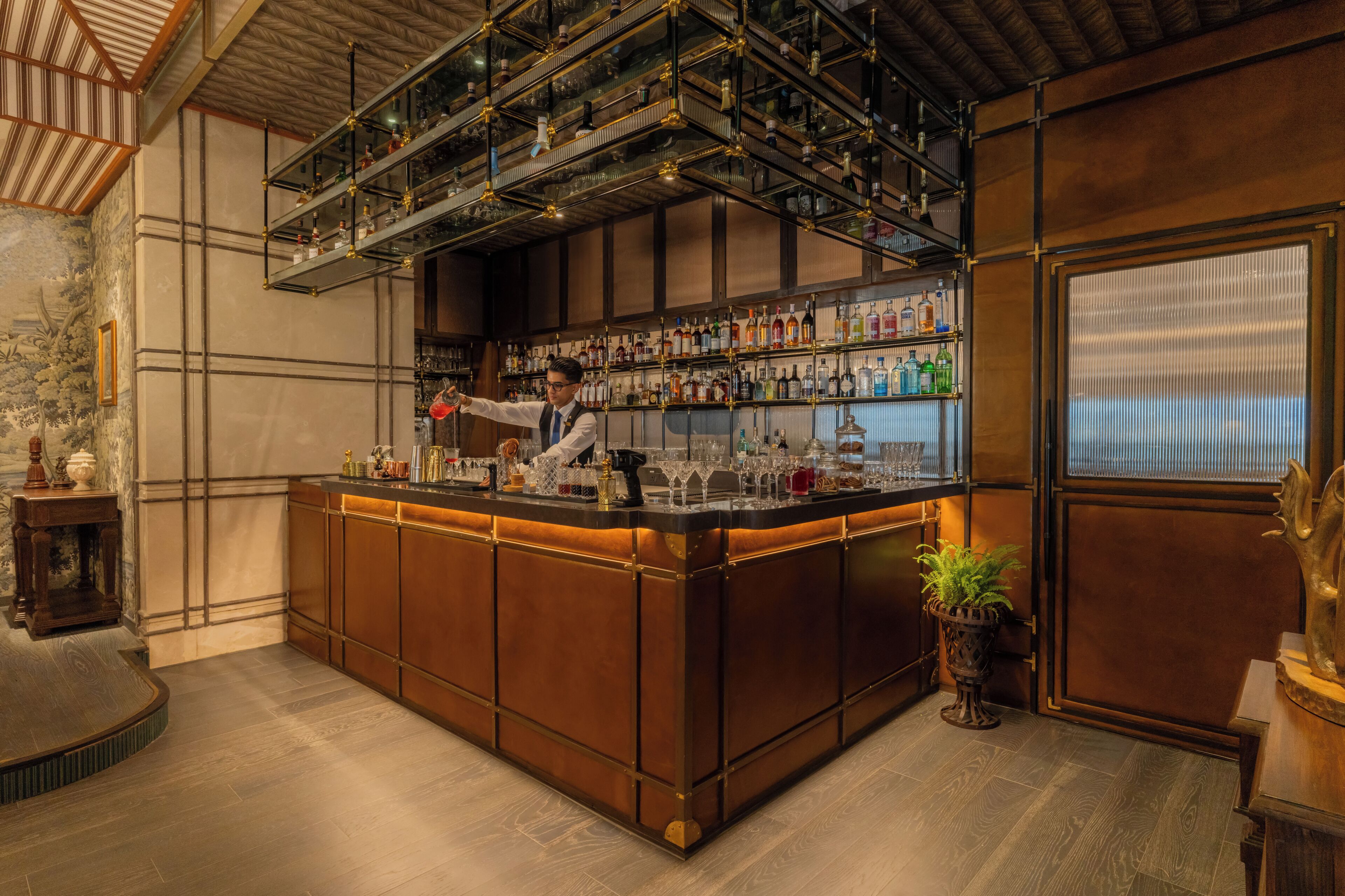 Uma área de bar com um bartender, prateleiras de garrafas de bebidas e uma planta decorativa.