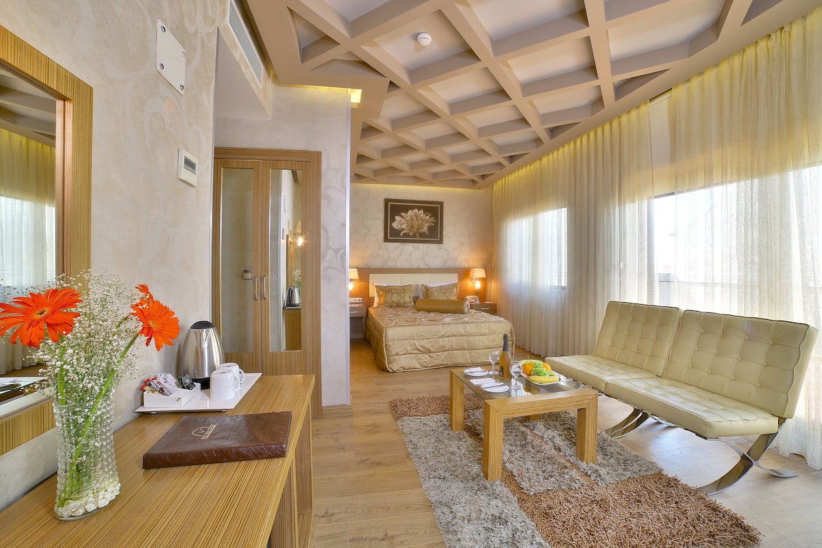 Quarto de hotel moderno com piso de madeira, sofá, mesa de jantar, cama e uma janela grande com cortinas.