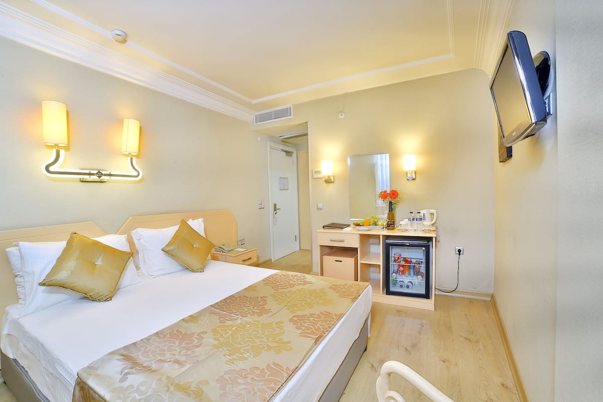 Quarto de hotel com cama, mesa de cabeceira, escrivaninha e uma pequena geladeira.