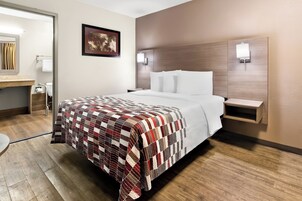 Quarto de hotel com uma cama grande, cabeceira de madeira, mesa de cabeceira, luminária fixada na parede e um quadro emoldurado.
