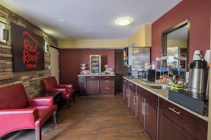 Um saguão de hotel com balcão de recepção, área de estar com cadeiras vermelhas e cozinha equipada com geladeira e cafeteira.