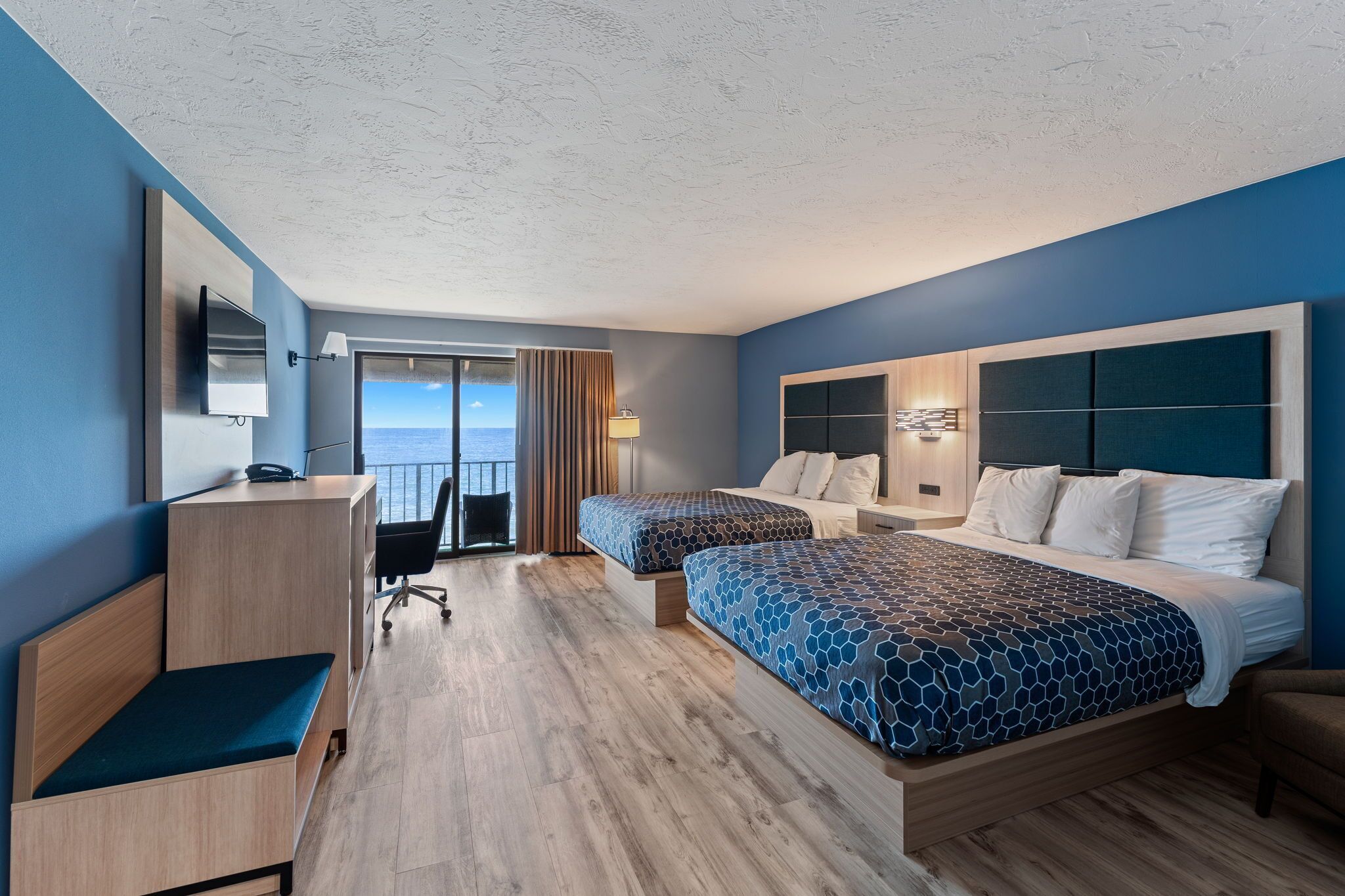 Quarto de hotel com duas camas, uma escrivaninha, uma cadeira e vista para o oceano.