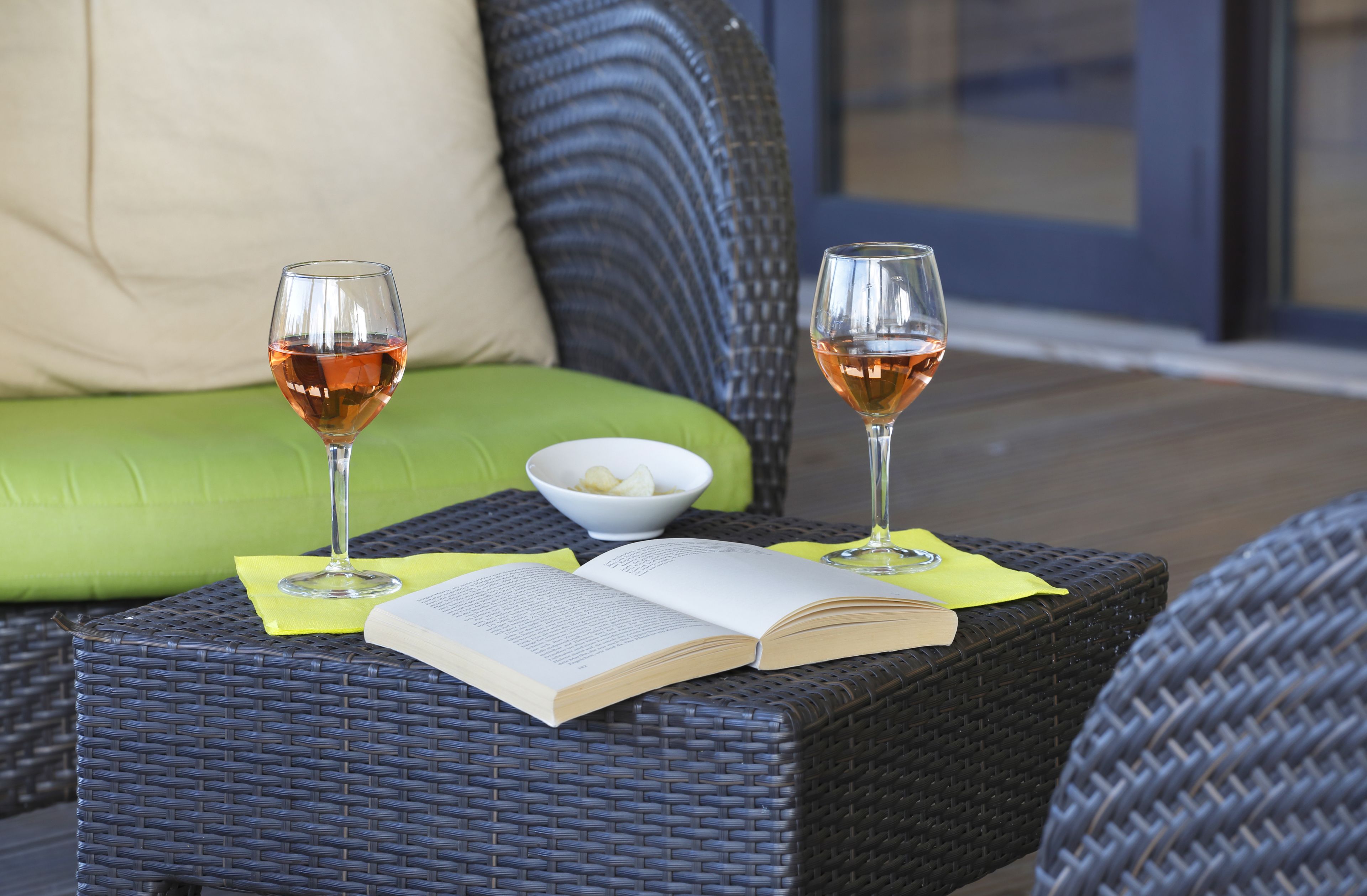 Uma mesa de vime com duas taças de vinho, uma tigela de petiscos e um livro aberto.
