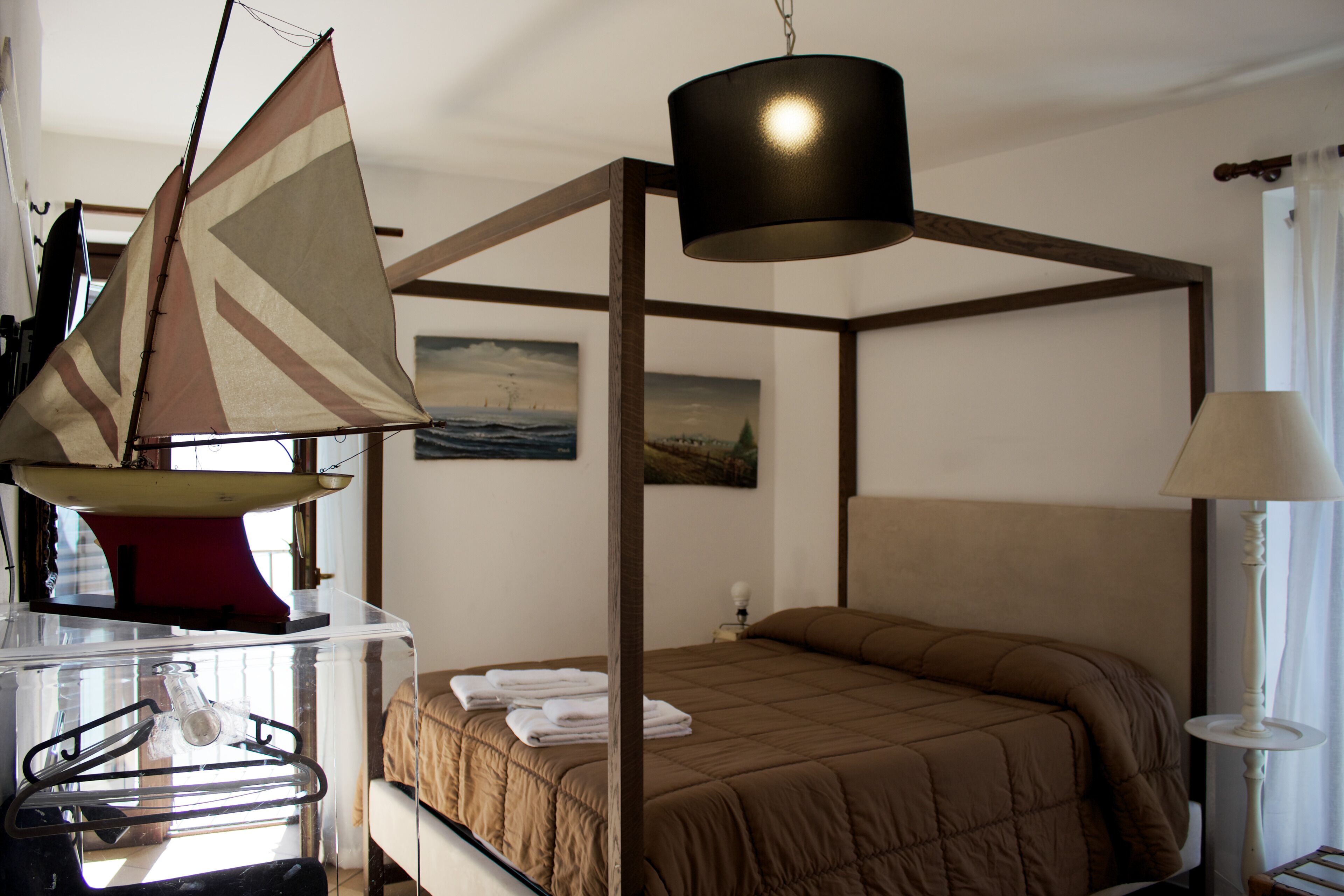 Uma cama de dossel com colcha marrom, uma mesa de vidro transparente com uma garrafa e um cabide, um abajur e uma decoração de veleiro.