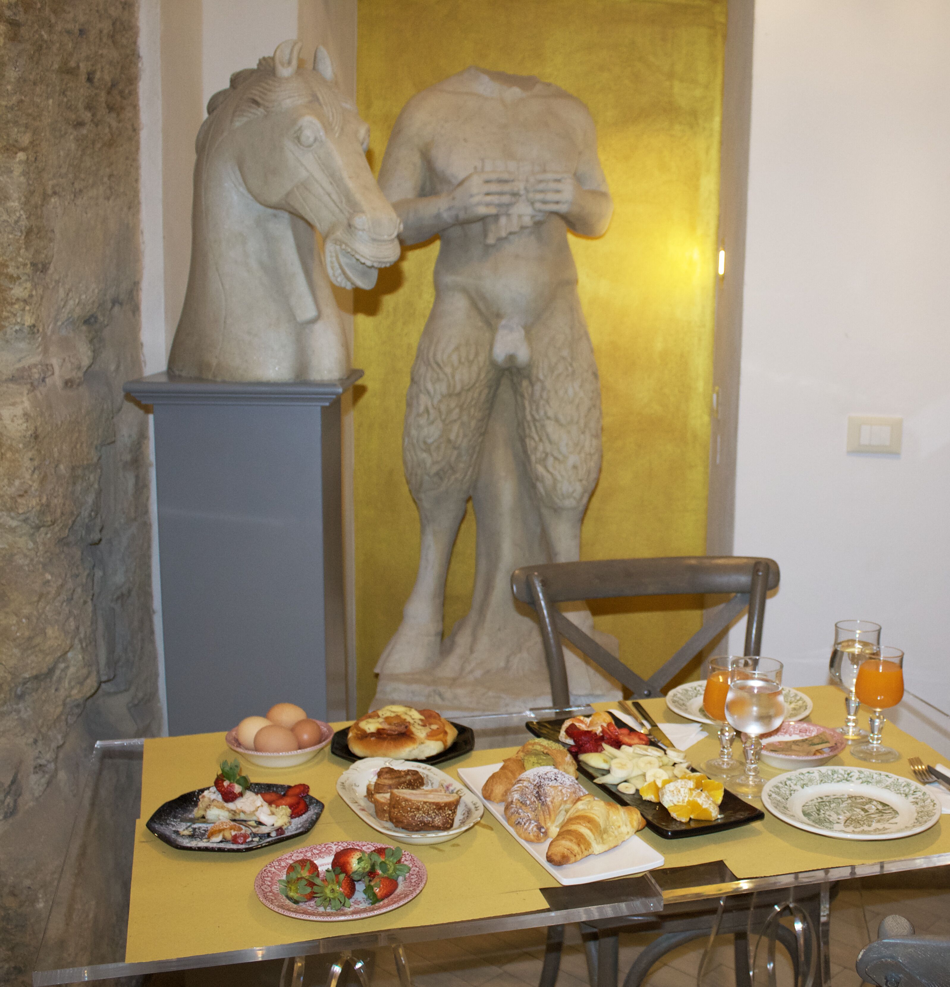 Uma mesa de jantar com diversos itens de café da manhã, uma estátua de um cavalo e uma figura humana, e uma cadeira.