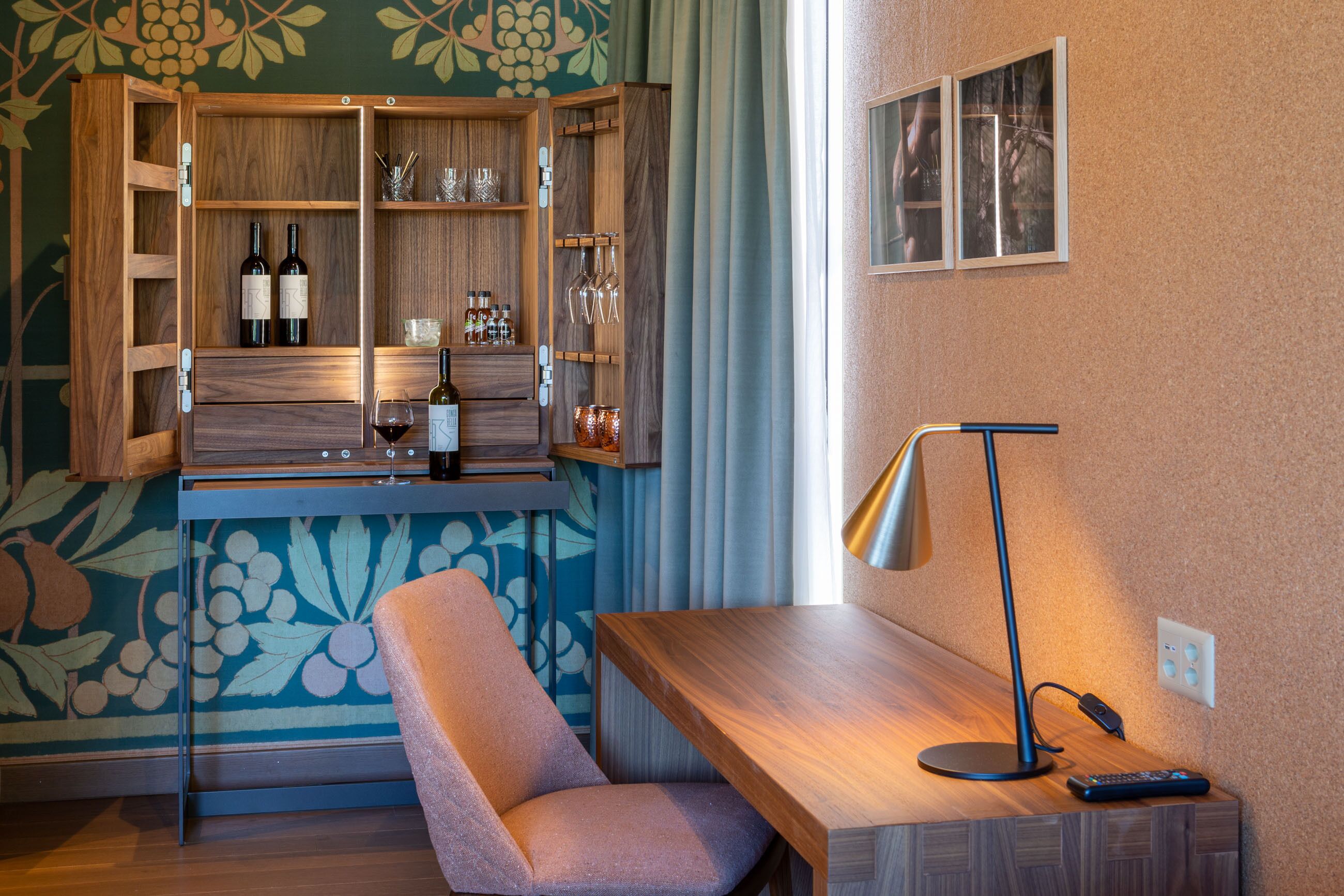 Um quarto com um armário de madeira com garrafas de vinho e copos, uma escrivaninha de madeira com uma luminária e uma cadeira.