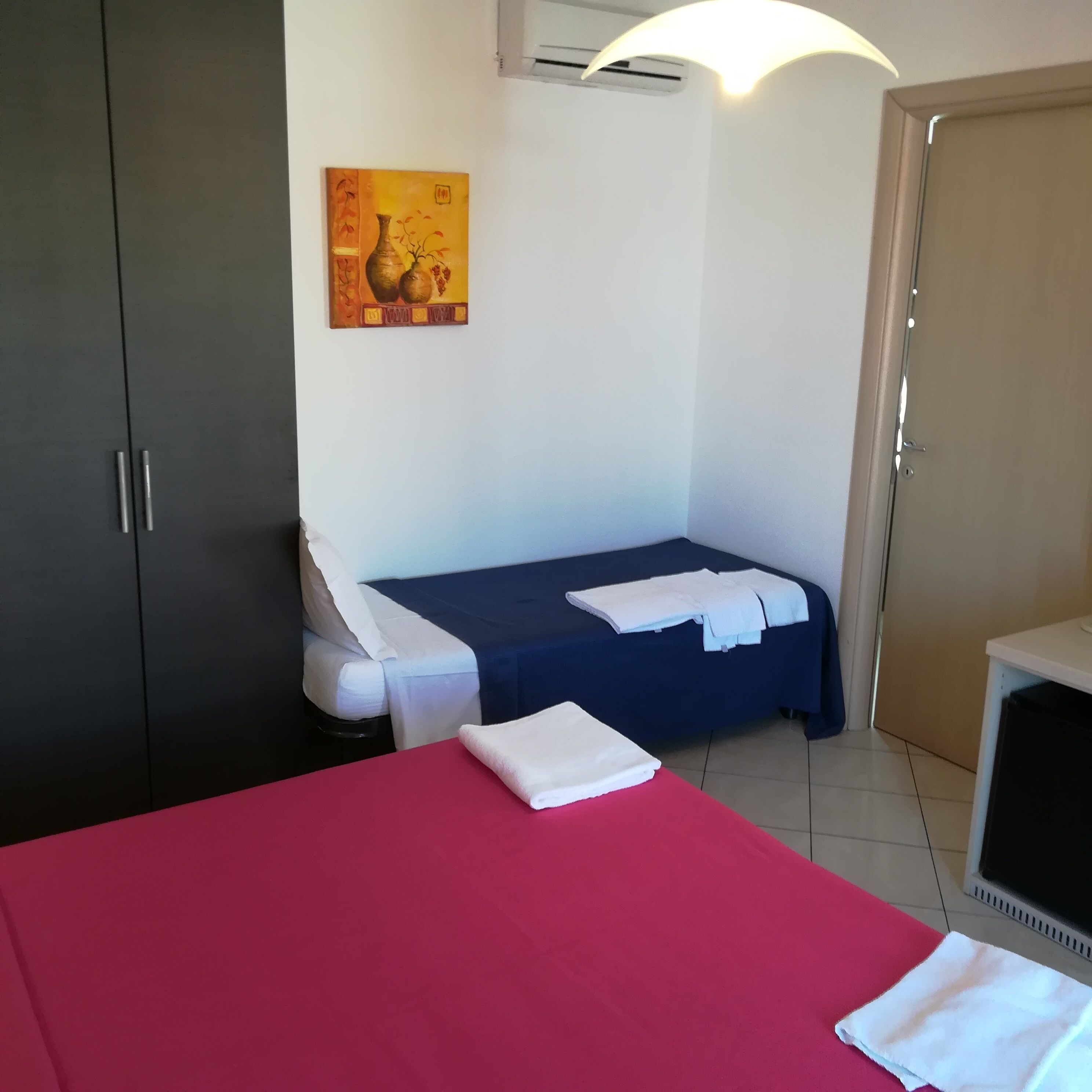 Quarto com uma cama vermelha, uma cama azul e um guarda-roupa. Há um quadro na parede e um ar condicionado sobre as camas.