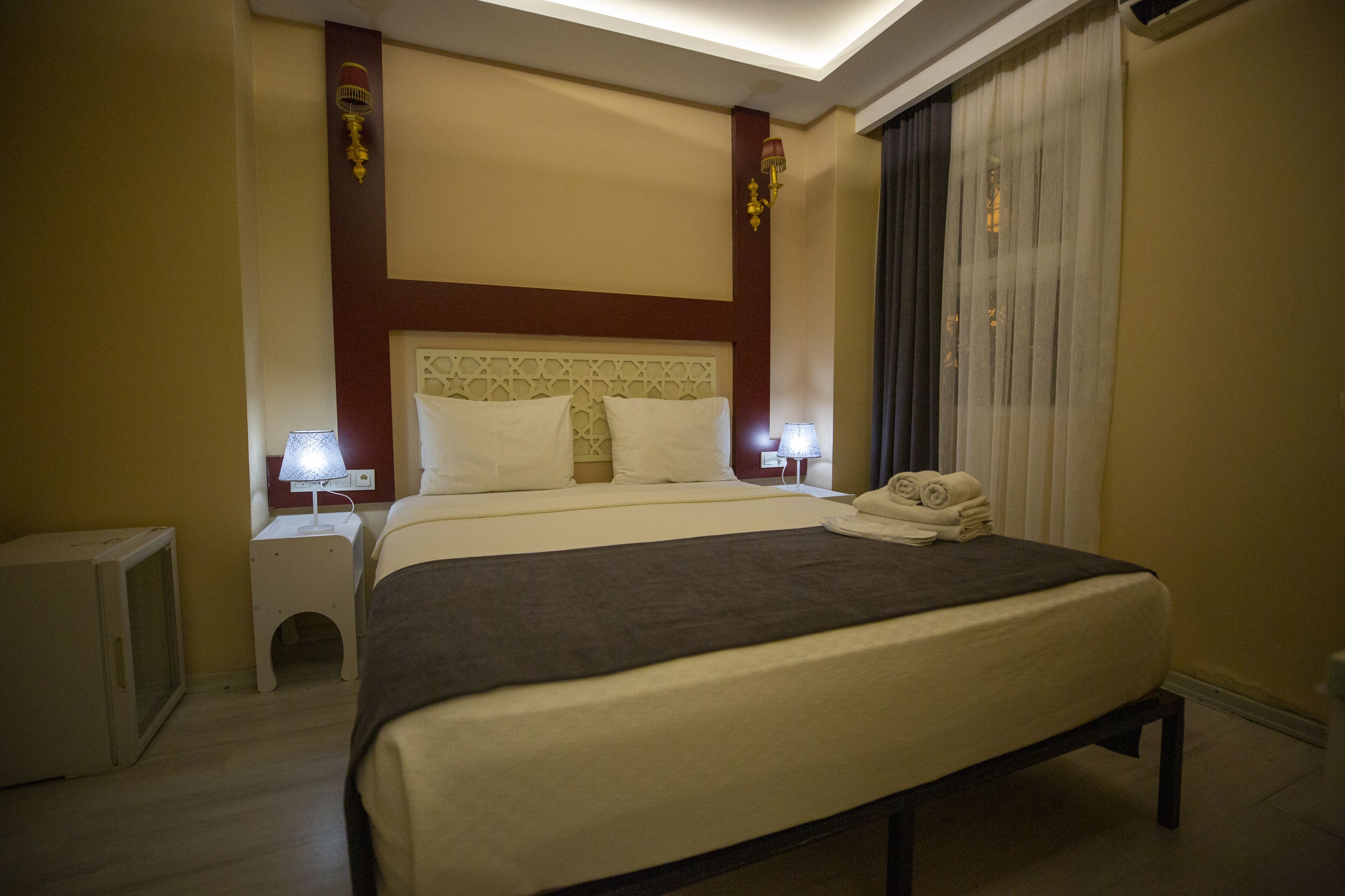Quarto de hotel com uma cama grande, duas mesas de cabeceira, abajur, telefone e janela com cortinas.