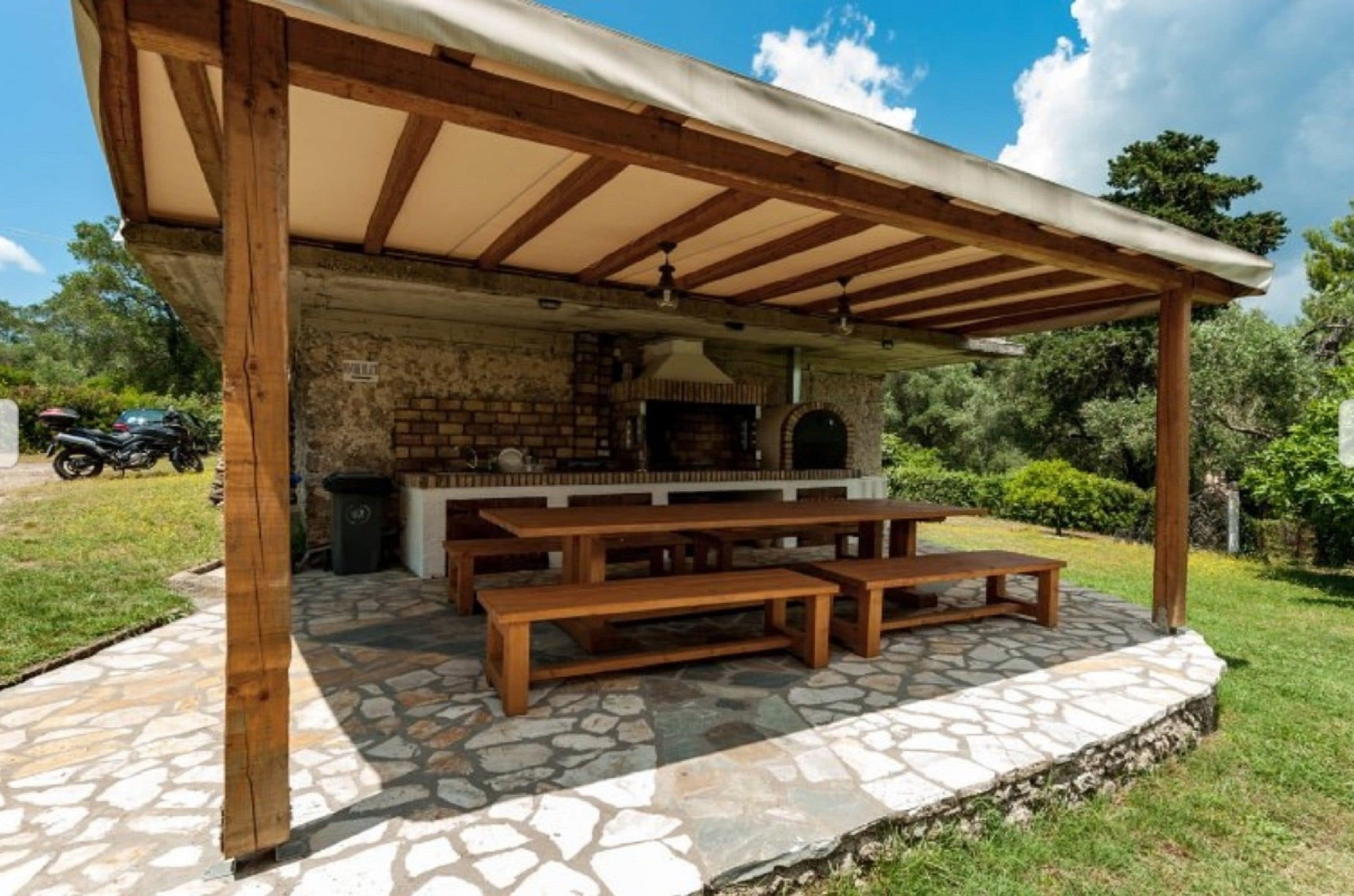 Uma área de churrasco ao ar livre com piso de pedra, bancos de madeira e forno de tijolos sob uma pérgola de madeira.