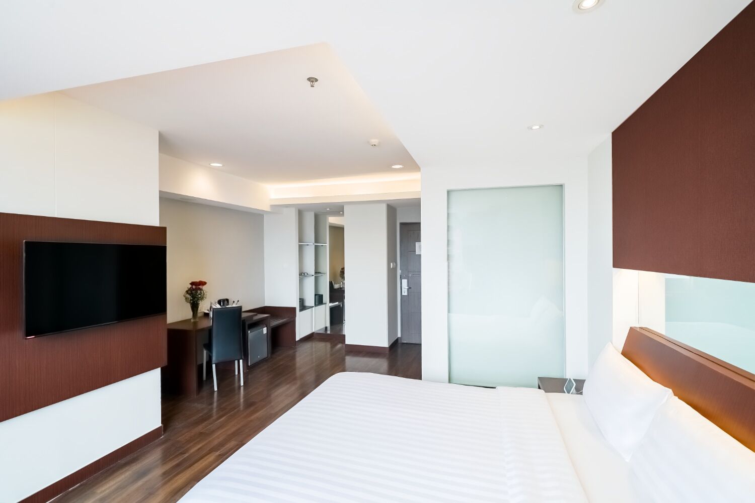 Quarto de hotel moderno com uma cama grande, uma televisão de tela plana na parede, uma escrivaninha com cadeira e uma porta de vidro que dá para outro cômodo.