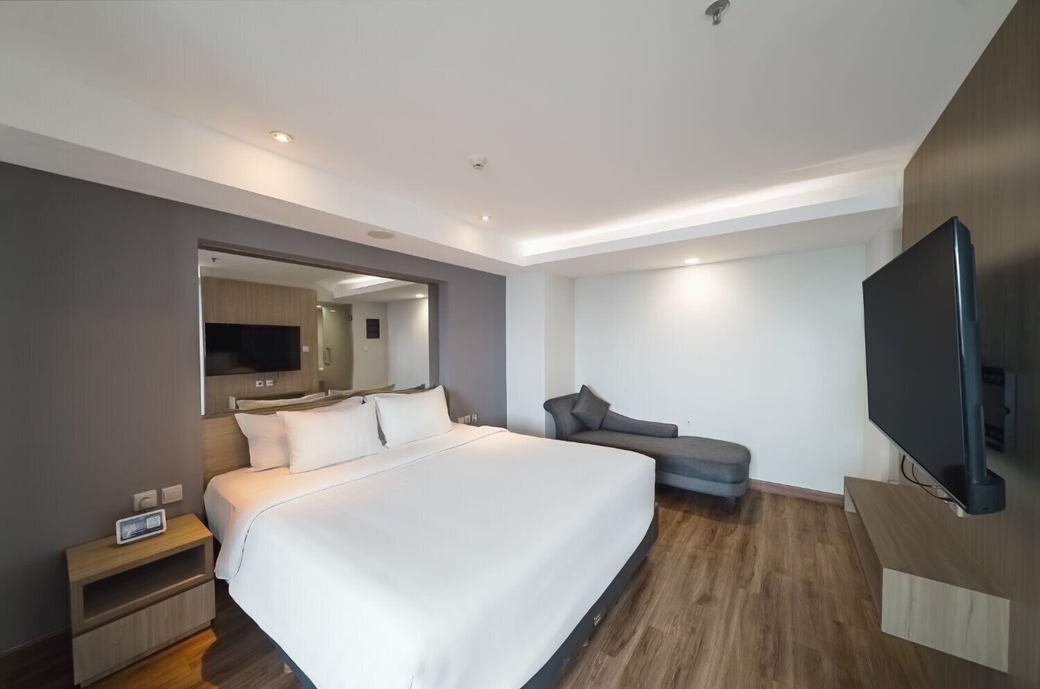 Quarto de hotel moderno com uma cama grande, televisão, mesa de cabeceira, sofá e espelho.